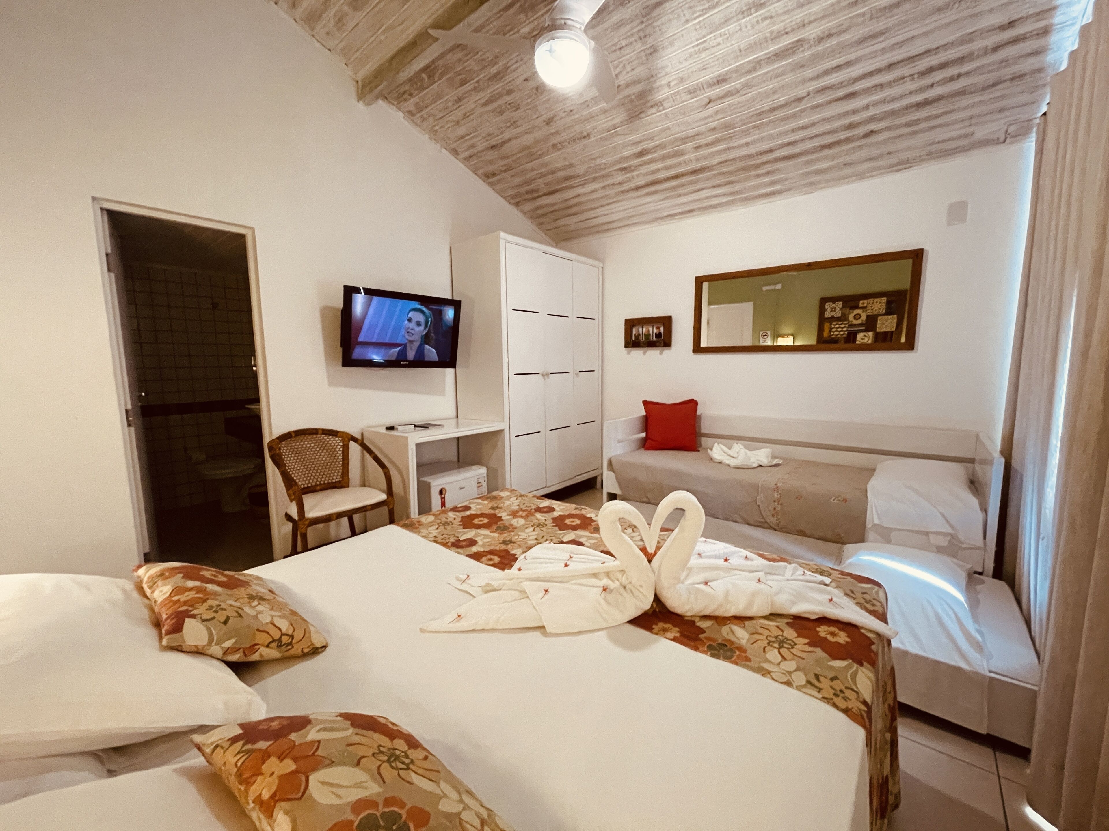 Quarto de hotel com cama, escrivaninha, televisão, guarda-roupa e banheiro visível por uma porta aberta.