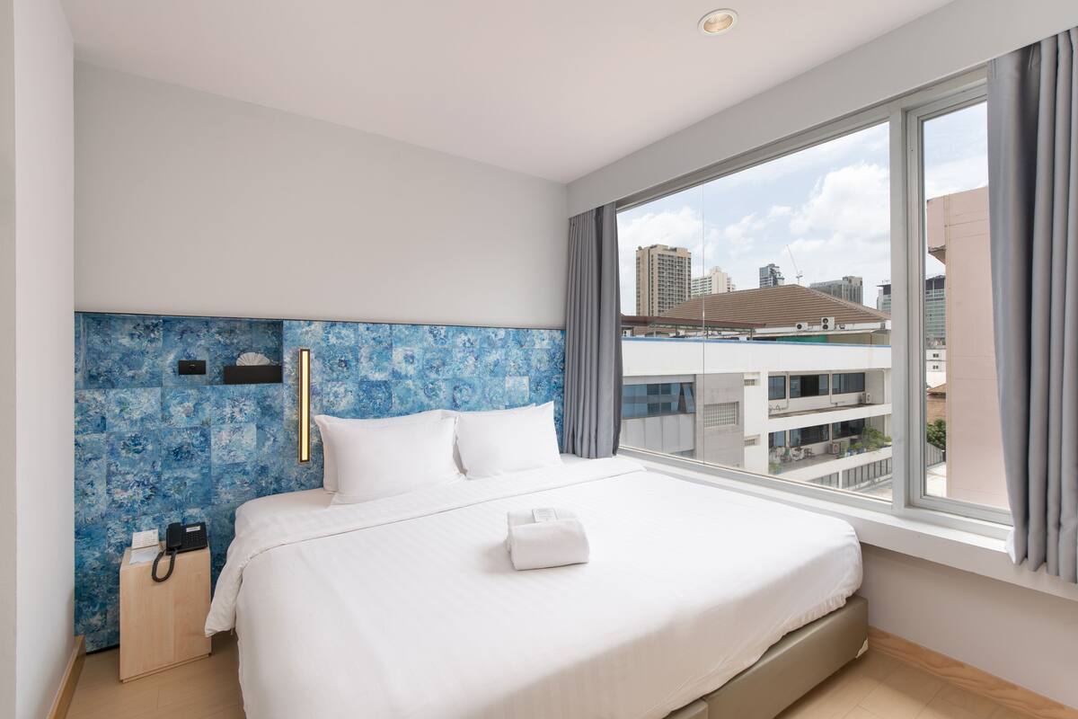 Quarto de hotel com uma cama grande, cabeceira de mosaico azul, um telefone e vista para edifícios pela janela.