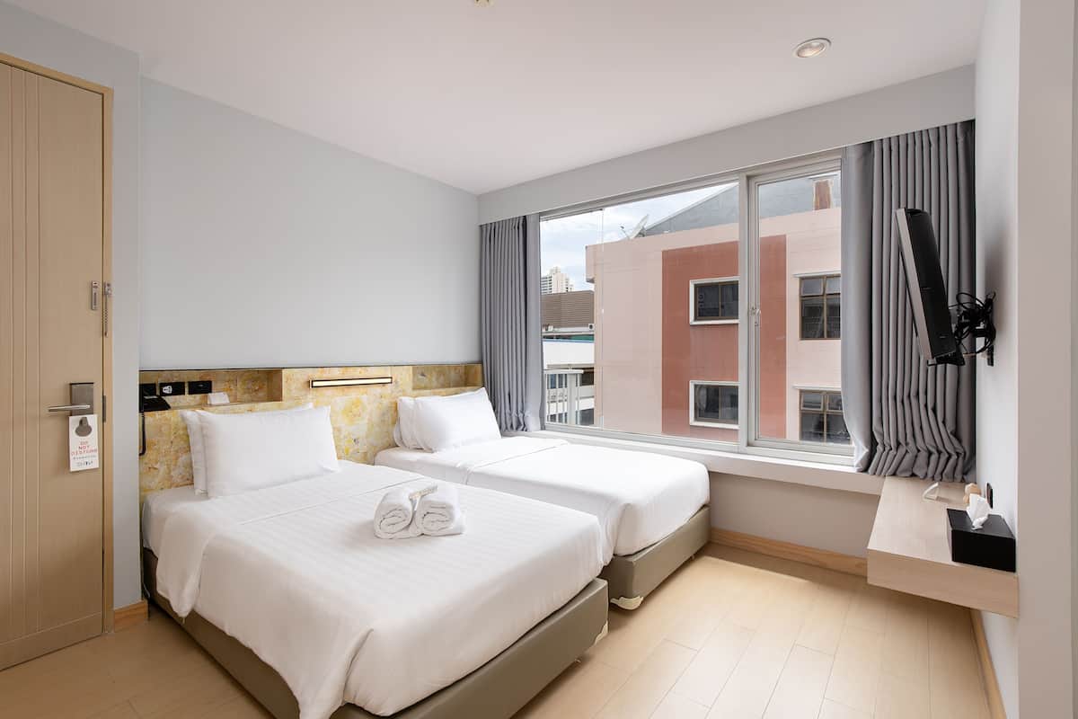 Quarto de hotel com duas camas, uma janela grande com cortinas, uma TV na parede e uma pequena mesa com abajur.