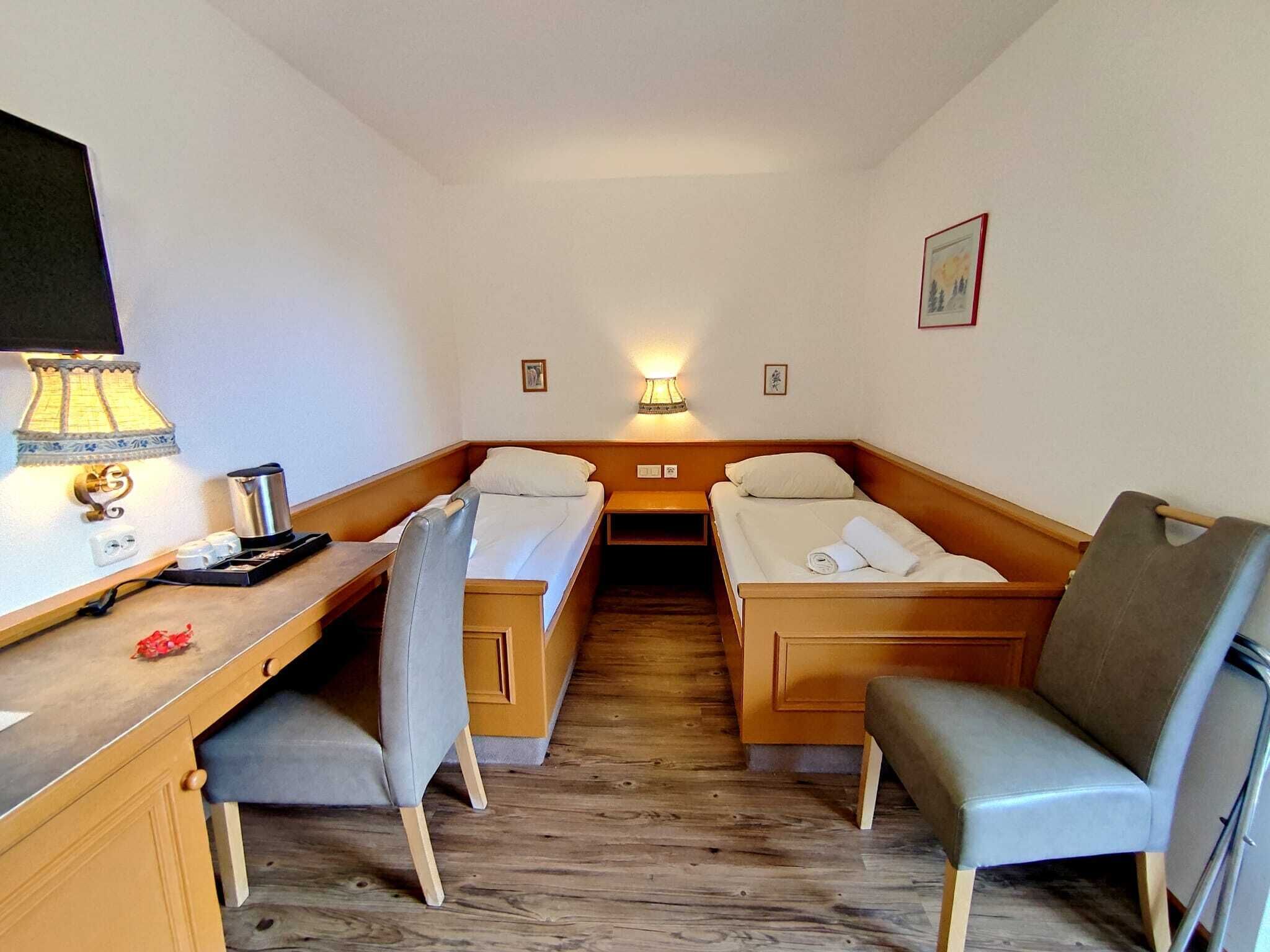 Quarto de hotel compacto com duas camas de solteiro, uma escrivaninha com luminária, uma cadeira e uma pequena mesa com chaleira.