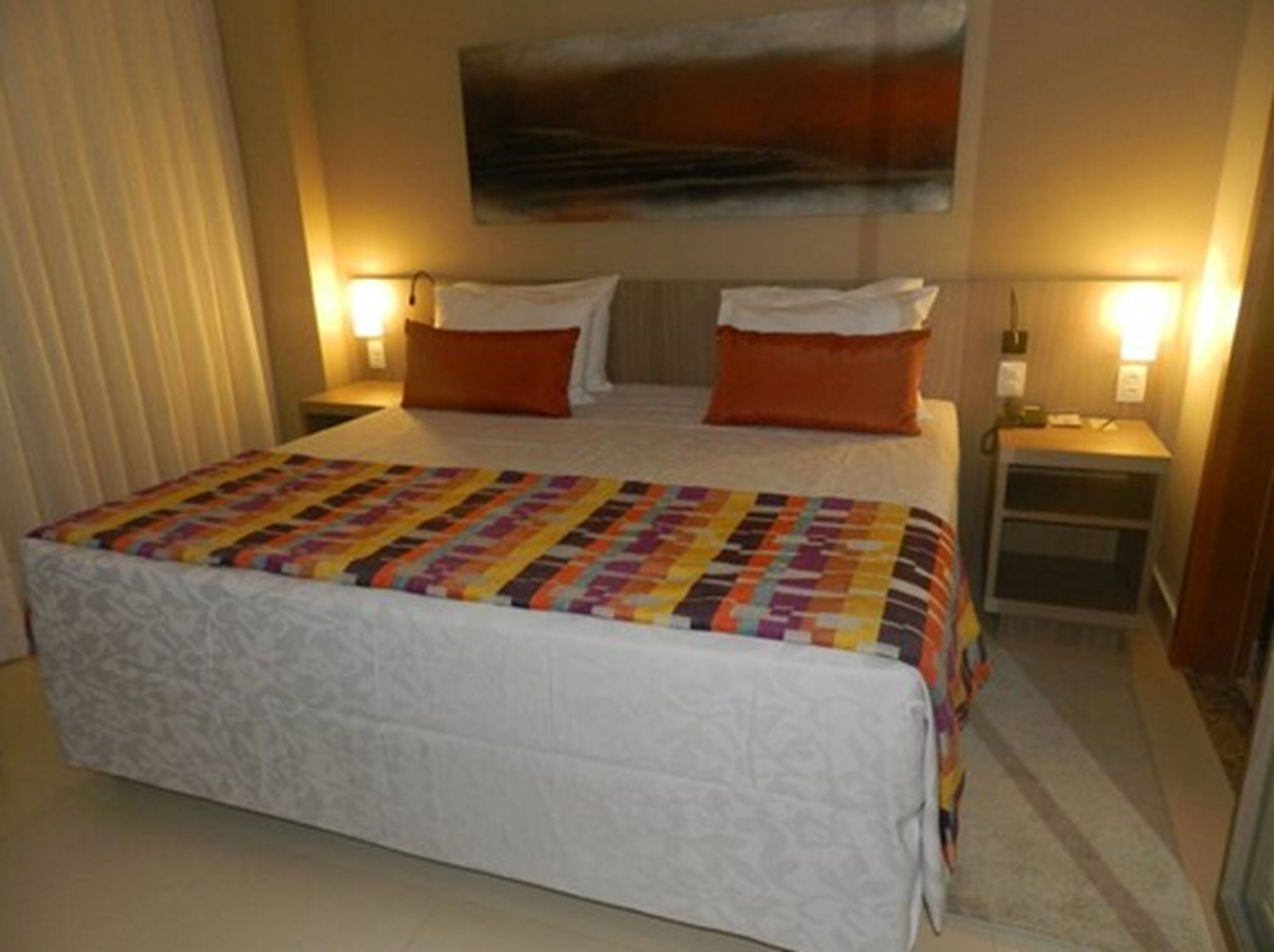 Quarto de hotel com uma cama, duas almofadas, uma mesa de cabeceira, um abajur e um quadro na parede.