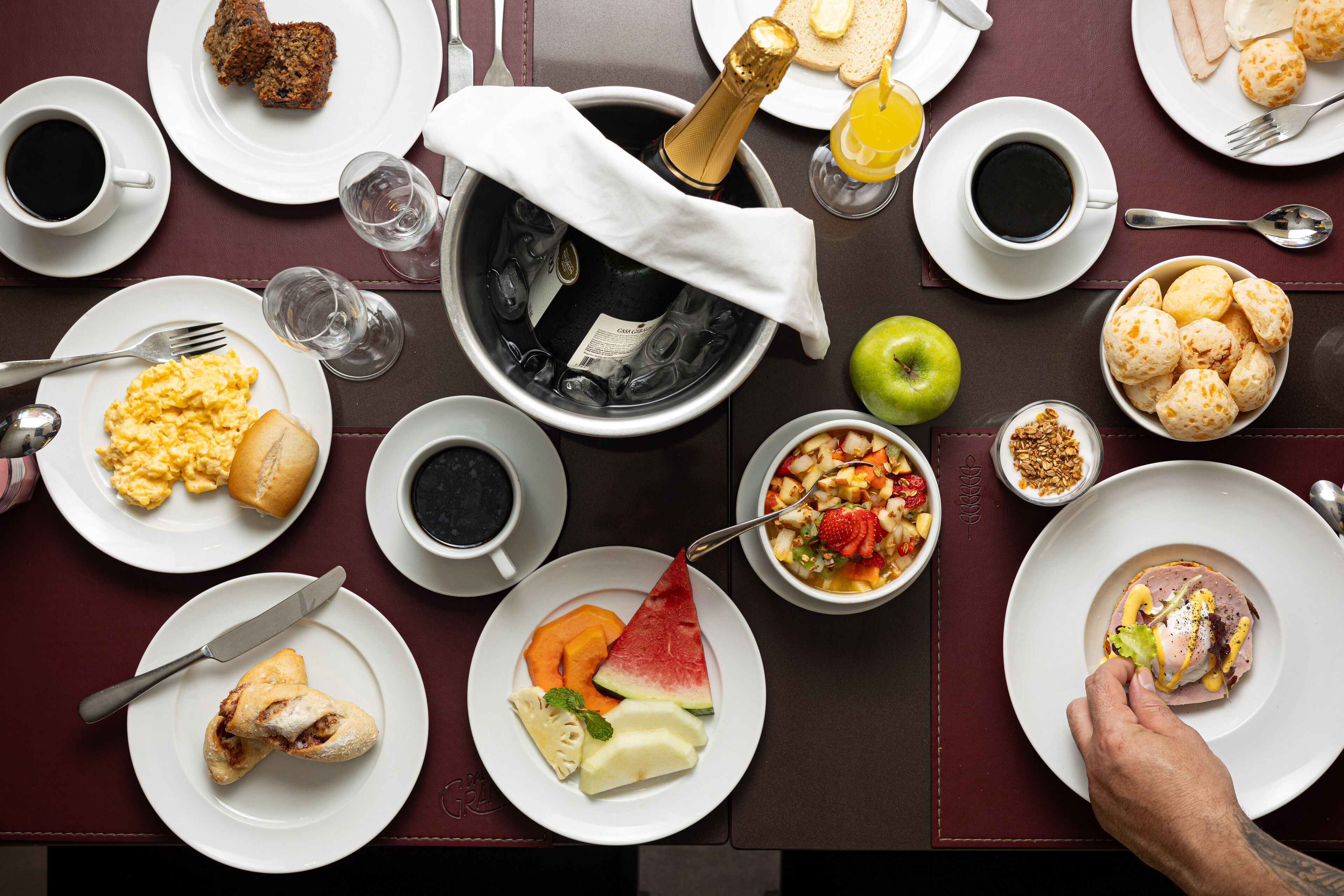 Mesa posta com diversos itens de café da manhã, incluindo ovos, torradas, frutas e bebidas.