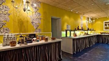 Disposição de buffet com parede amarela e papel de parede floral, um balcão comprido com diversos alimentos e um candelabro.