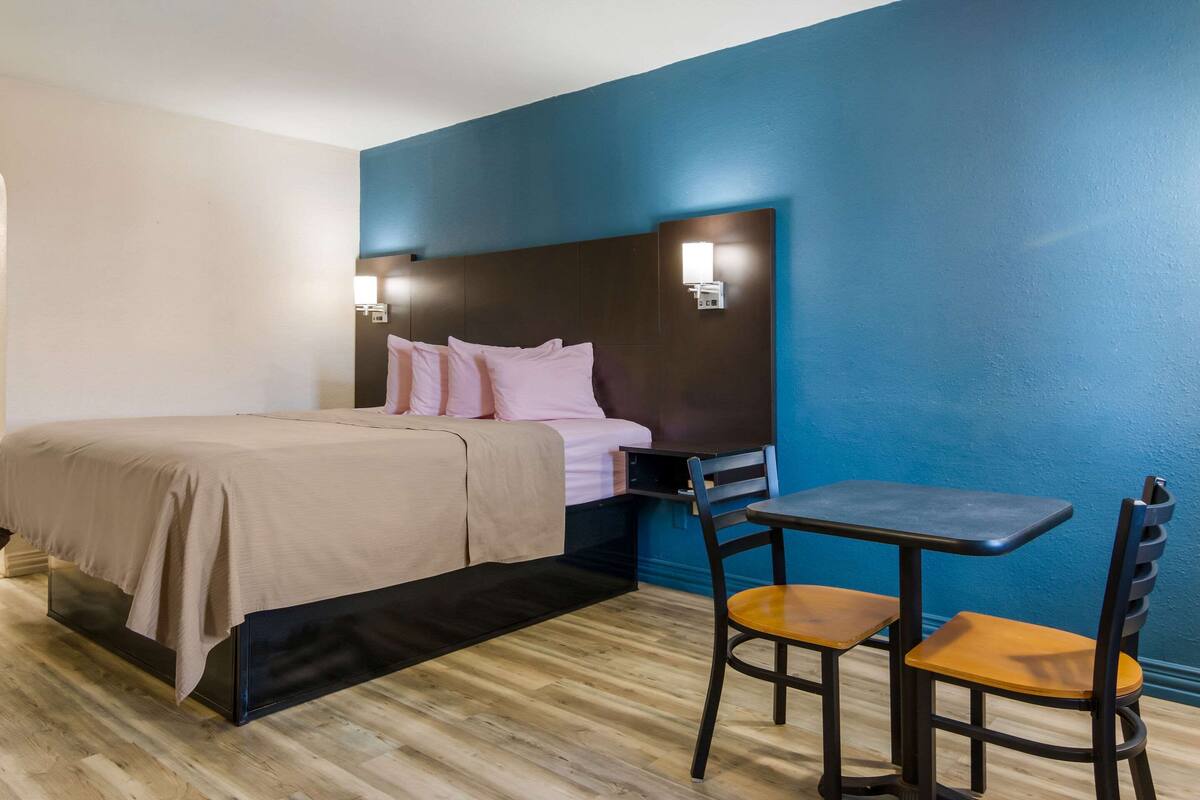 Quarto de hotel com cama, escrivaninha, duas cadeiras e uma parede azul.