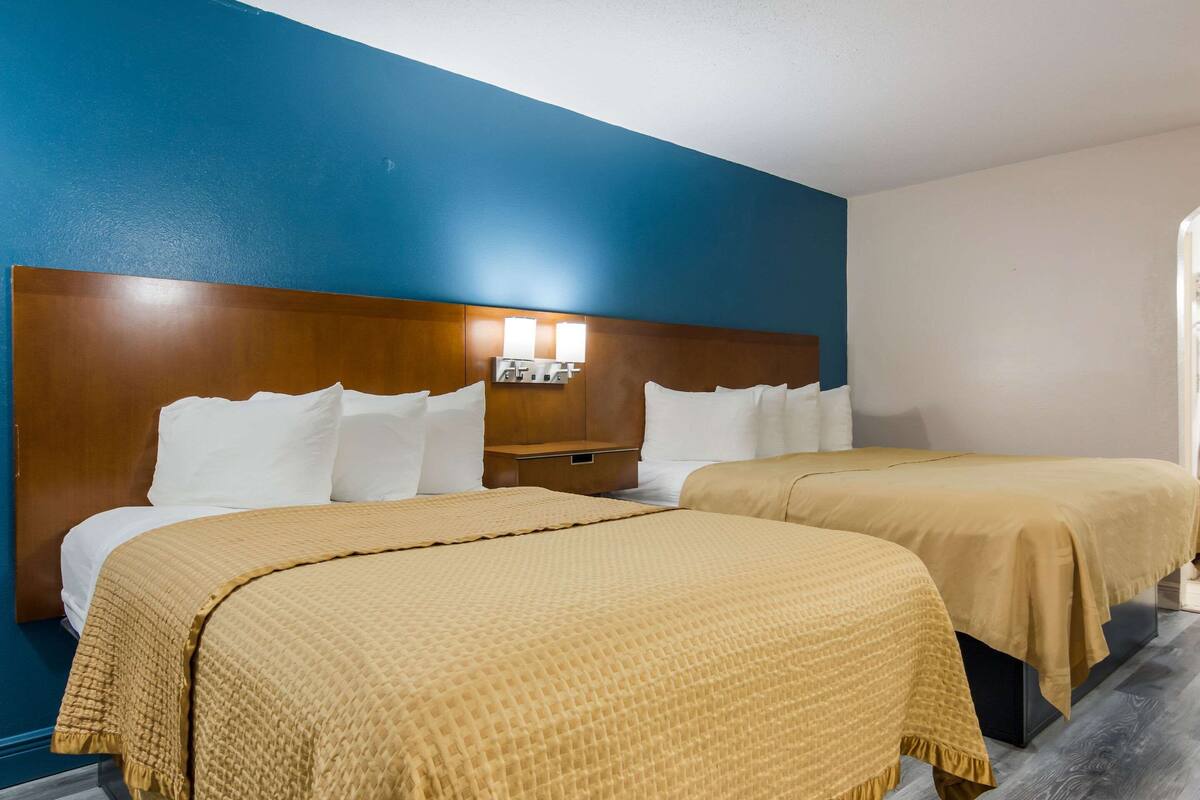 Quarto de hotel com duas camas, cabeceira de madeira, mesa de cabeceira com abajur e parede azul decorativa.