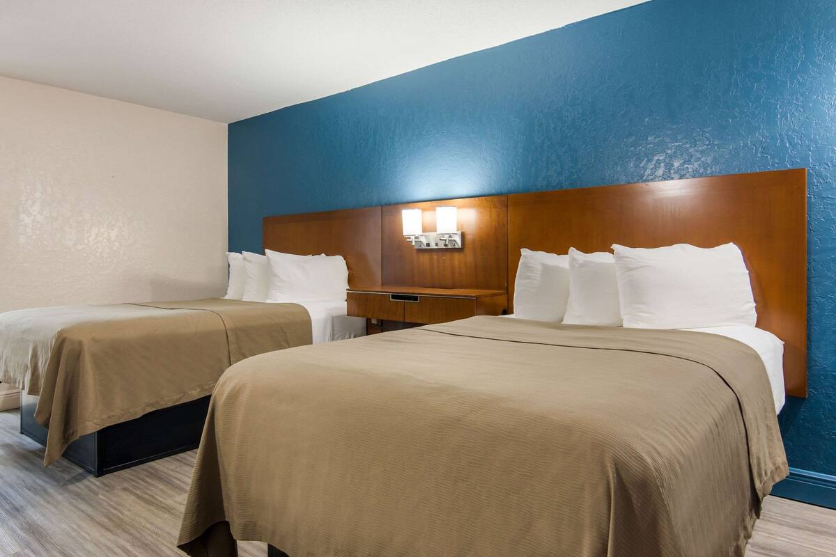 Quarto de hotel com duas camas, cabeceira de madeira, mesa de cabeceira e luminária fixa na parede.