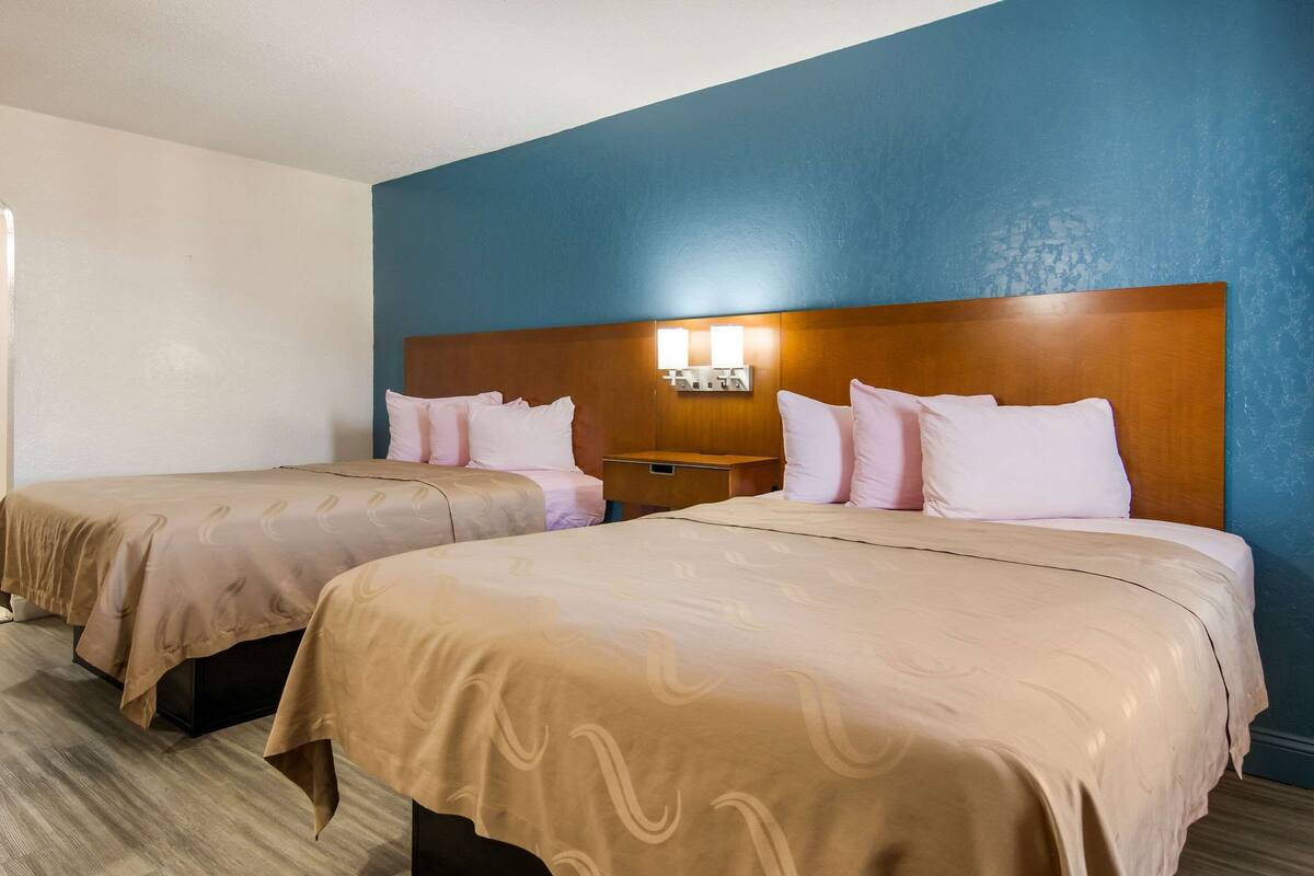 Quarto de hotel com duas camas, uma mesinha de cabeceira e um candeeiro fixo na parede.
