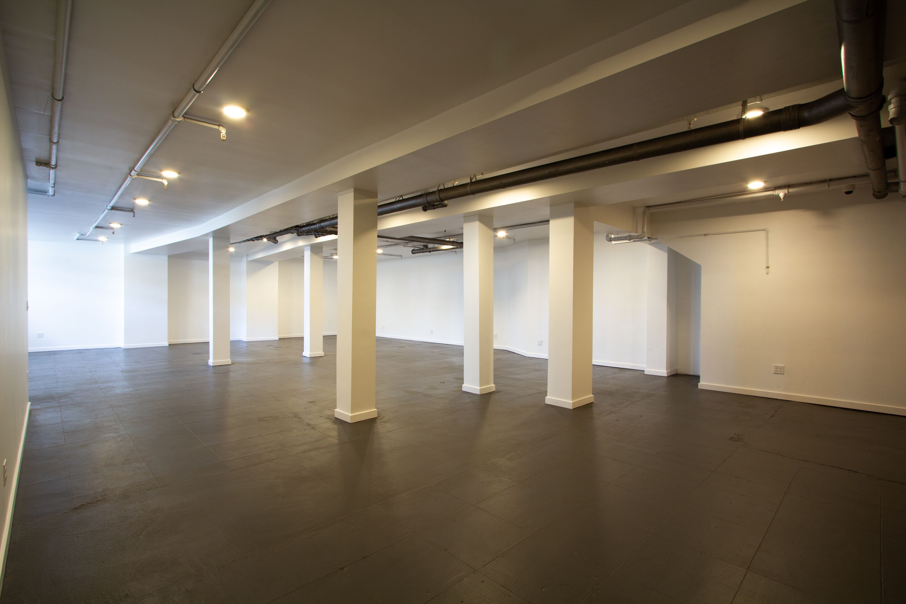 Um espaço amplo e vazio, com pilares brancos e chão escuro, apresentando tubulações aparentes e um design limpo e minimalista.