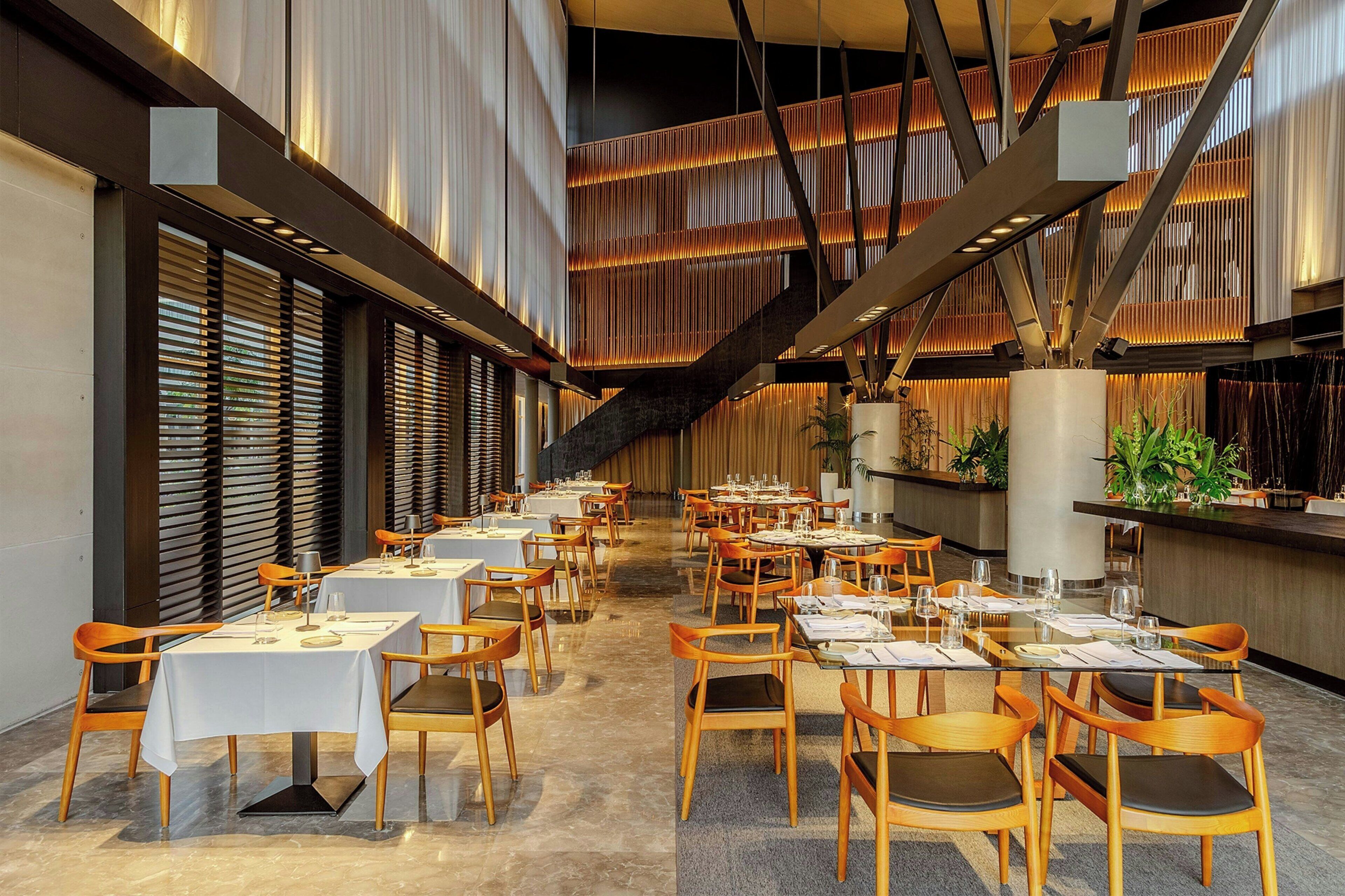 Uma ampla área de jantar com várias mesas e cadeiras, uma escadaria central e design de interiores moderno.