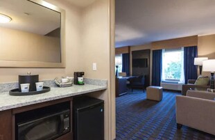 Quarto de hotel com cozinha compacta, um espelho grande, televisão de tela plana, área de estar com sofá e poltronas, e janela com cortinas.