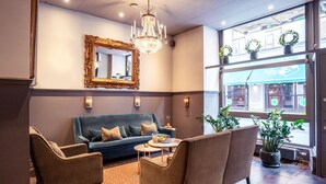 Um hall com um sofá azul, duas poltronas, um candelabro, um espelho e uma janela com plantas.