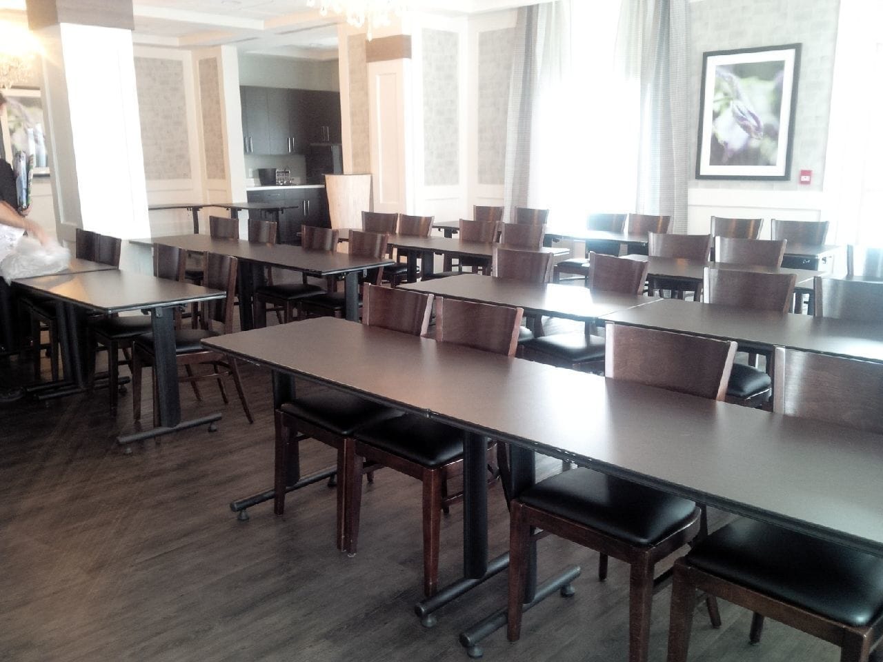 Uma sala de conferências com várias mesas e cadeiras, uma área de cozinha ao fundo e um quadro pendurado na parede.