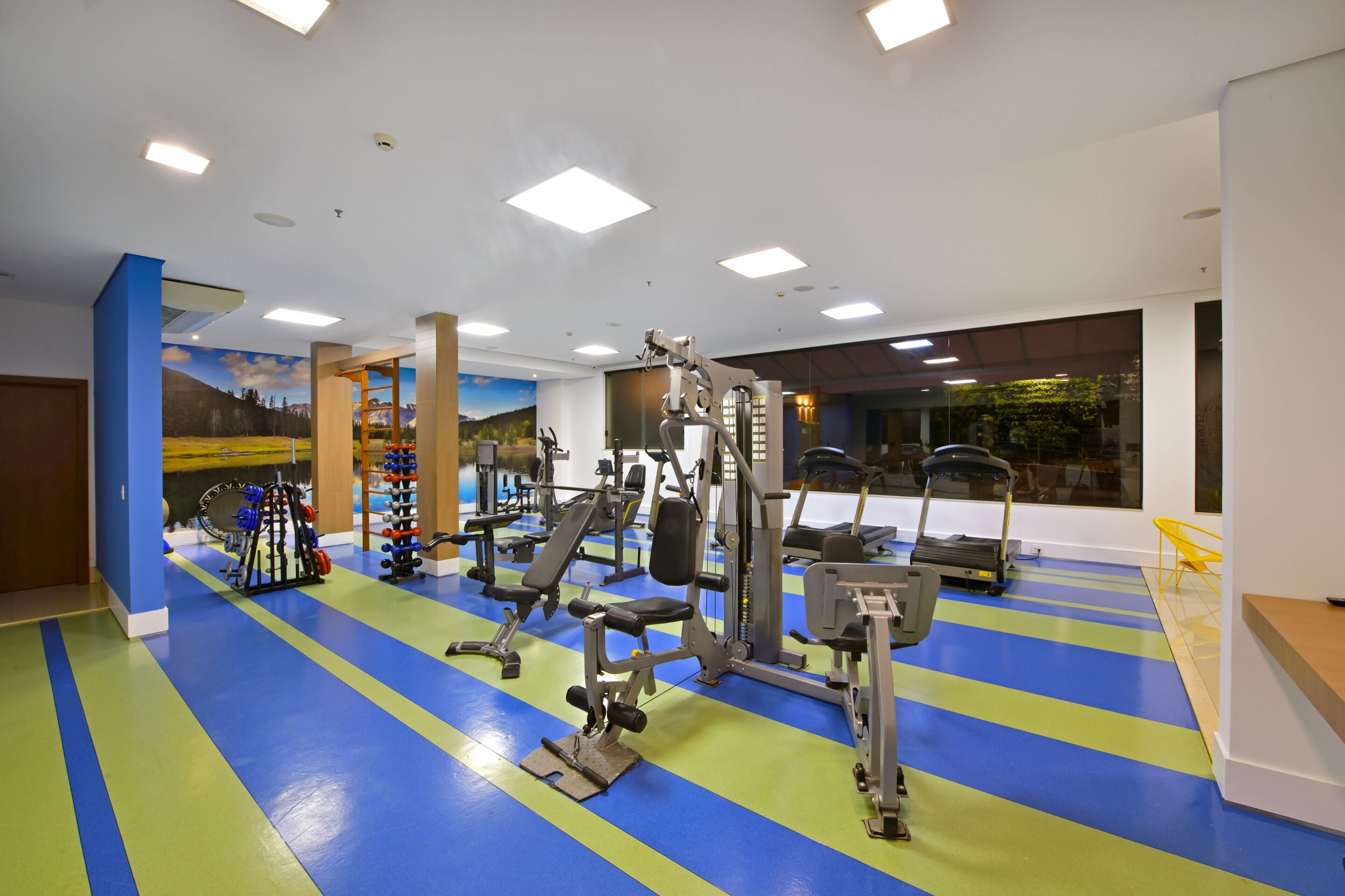 Uma academia com diversos equipamentos de exercício, piso azul e verde, e grandes janelas com vista para áreas verdes.