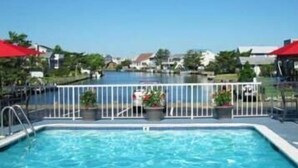 Piscina retangular com deck branco e grade de metal, cercada por plantas em vasos e guarda-sóis vermelhos.