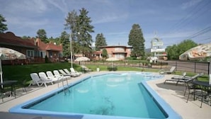 Uma piscina retangular com espreguiçadeiras e guarda-sóis ao redor, cercada por edifícios e árvores.