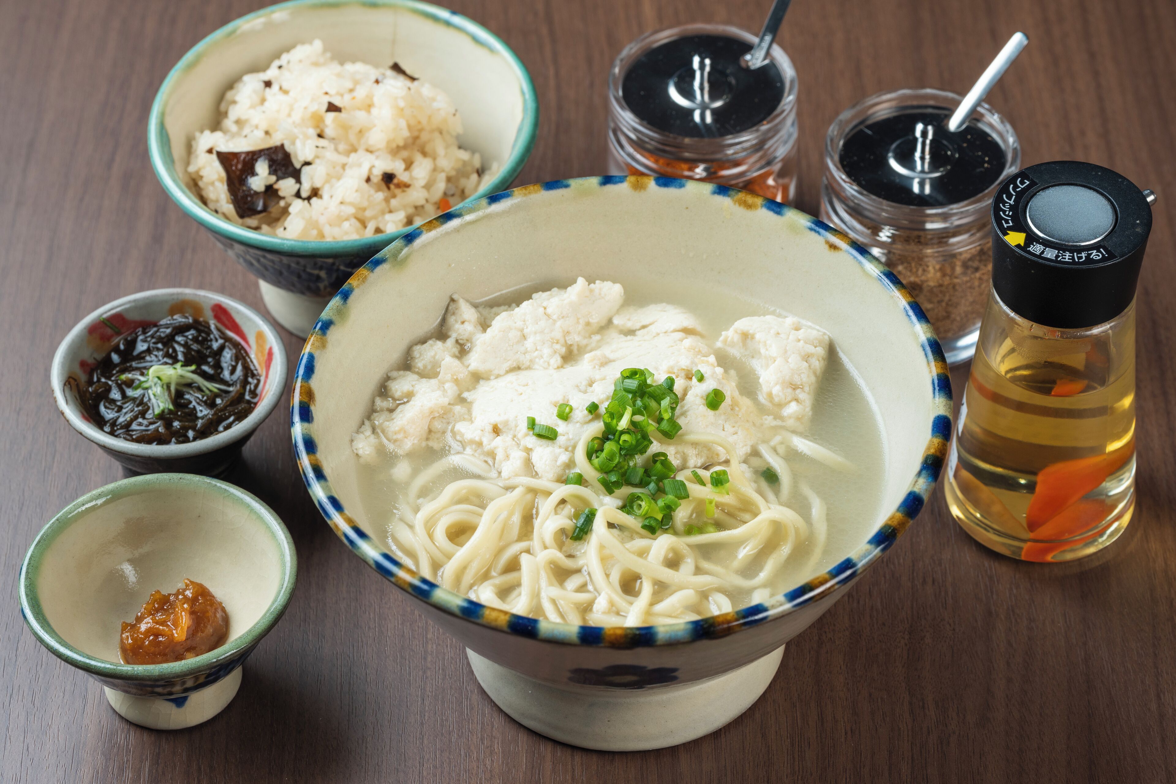 Uma tigela de ramen com macarrão, frango e cebolinha, acompanhada de pequenas tigelas com acompanhamentos e uma garrafa de molho de soja.