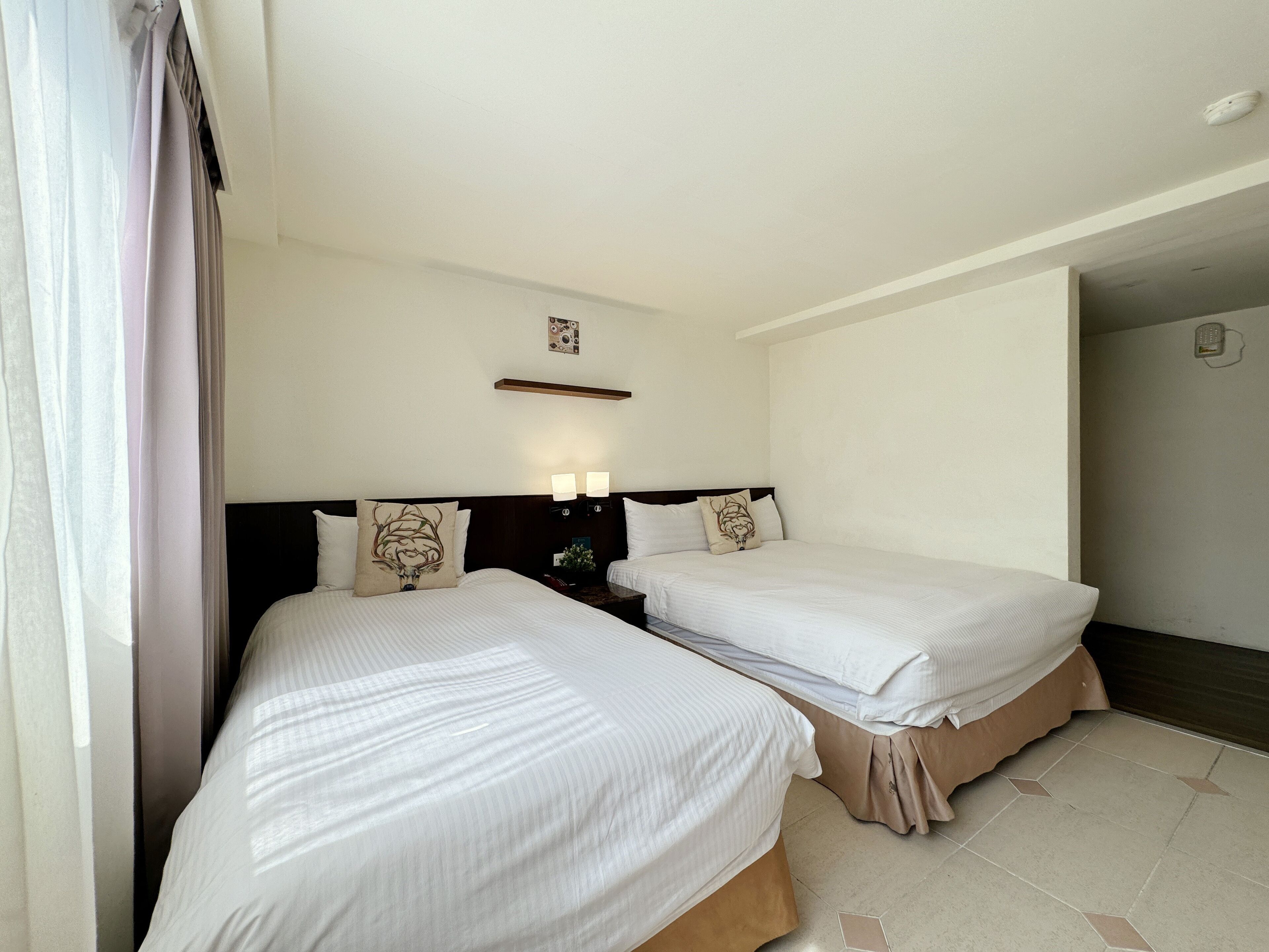 Quarto de hotel com duas camas individuais, uma mesa de cabeceira com abajur, uma pequena prateleira sobre a cama e uma parede lisa com interruptor de luz e detector de fumaça.