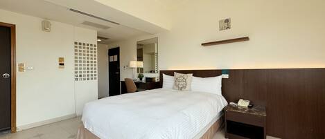 Quarto de hotel com uma cama grande, uma mesa de cabeceira com abajur, cabeceira de madeira e uma pequena prateleira.