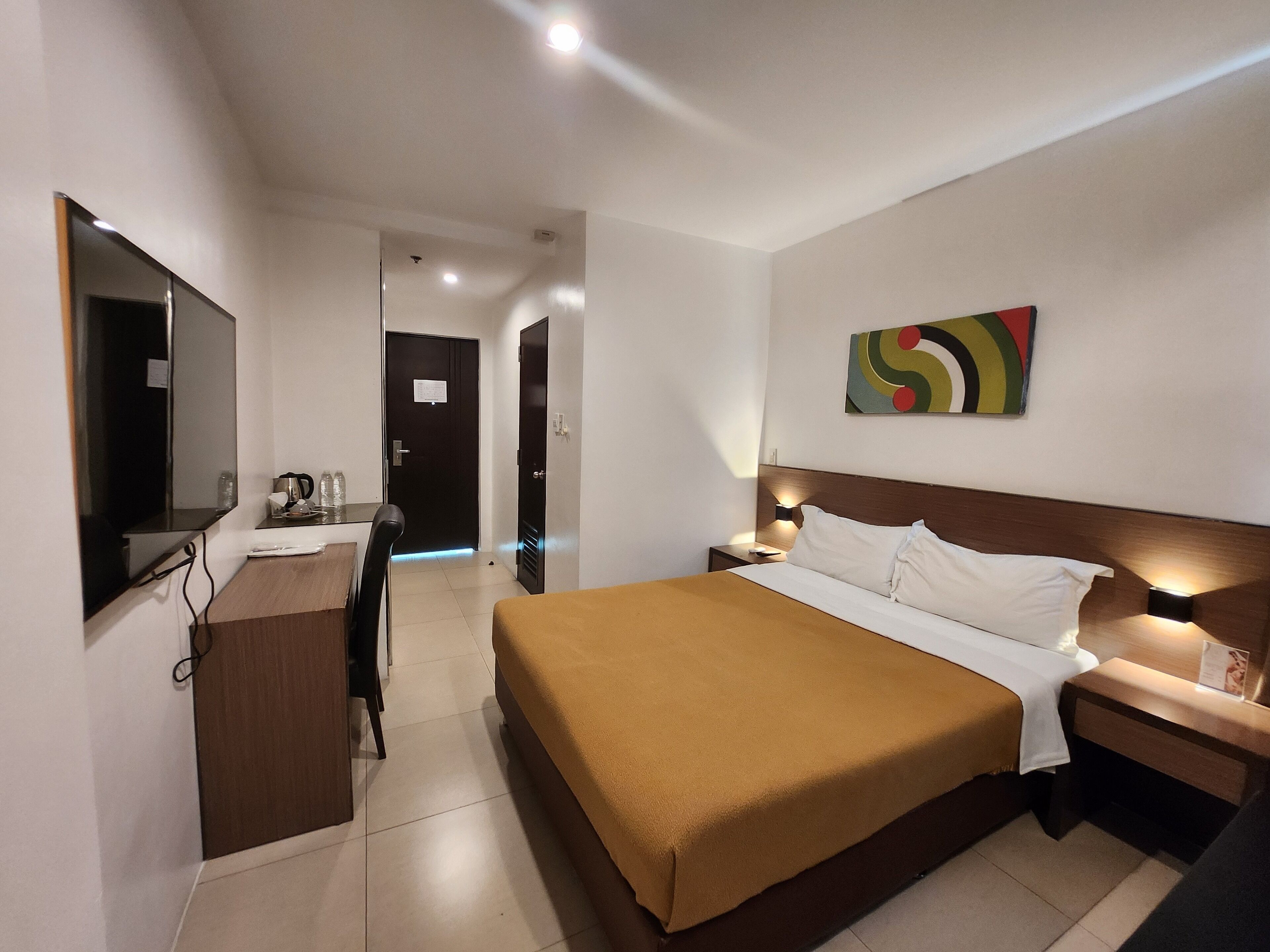 Quarto de hotel com cama, mesa de cabeceira, escrivaninha e televisão.