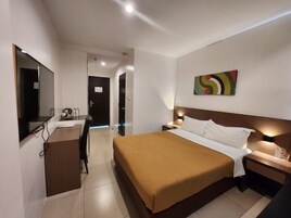 Quarto de hotel com cama, mesa de cabeceira, escrivaninha e televisão.
