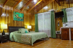 Um quarto com painéis de madeira, uma cama, duas mesas de cabeceira com luminárias, uma televisão montada na parede e uma pequena área de kitchenette.