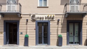 Entrada de hotel com uma placa indicando "Cuneo Hotel". Há duas portas, cada uma com um pequeno vaso de plantas em frente. O edifício tem fachada bege e uma varanda com grades decorativas.