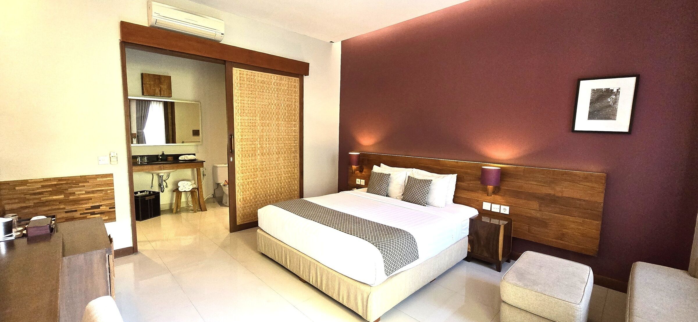 Quarto de hotel com cama, abajures, escrivaninha, cadeira e banheiro visível por uma porta aberta.