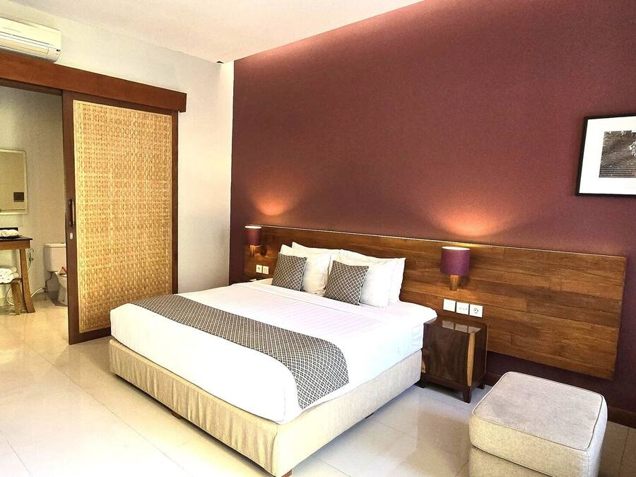 Quarto de hotel com cama, abajures, escrivaninha, cadeira e banheiro visível por uma porta aberta.