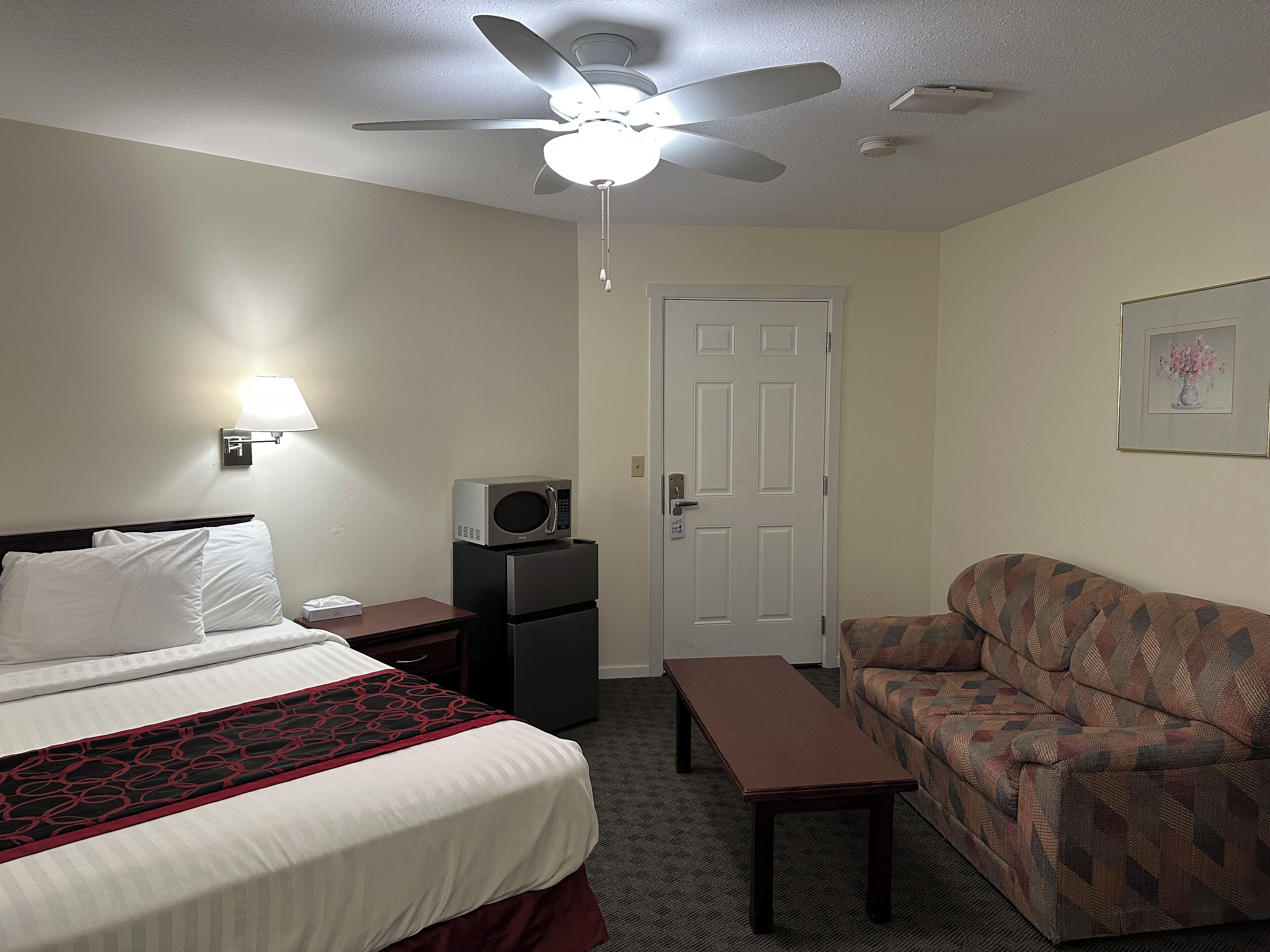 Quarto de hotel com cama, escrivaninha, micro-ondas, frigorífico, sofá, ventoinha no teto e porta.
