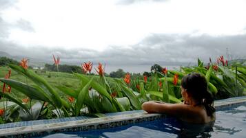 Mulher relaxando em uma piscina, com vista para campos verdes e flores vermelhas.