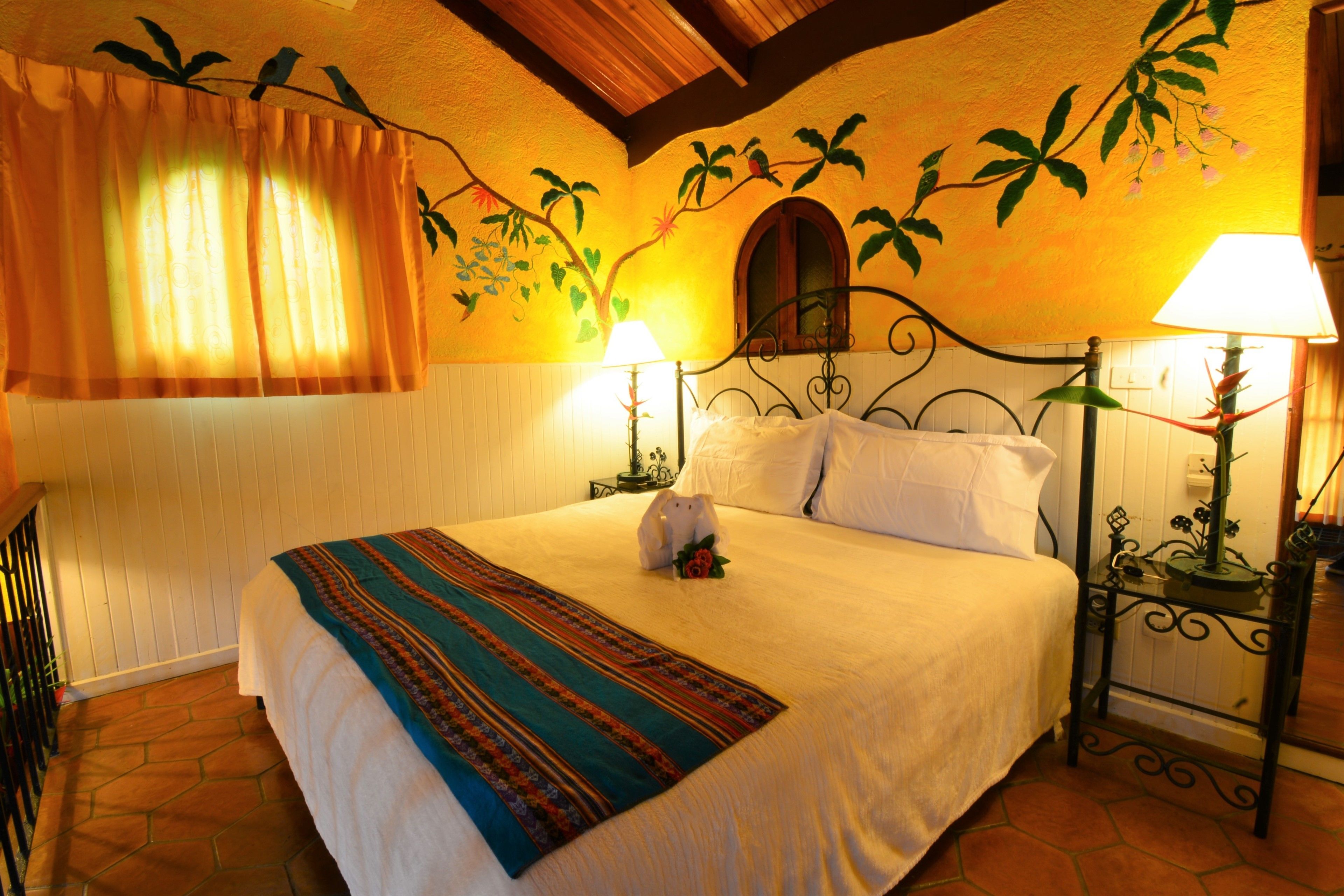 Uma cama bem arrumada com cabeceira decorativa, duas mesas de cabeceira com abajures e uma janela com cortinas.