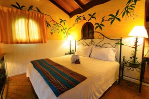 Uma cama bem arrumada com cabeceira decorativa, duas mesas de cabeceira com abajures e uma janela com cortinas.