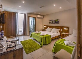 Quarto de hotel com duas camas, uma escrivaninha com cadeira, uma mesinha com uma garrafa de água e um tapete verde.