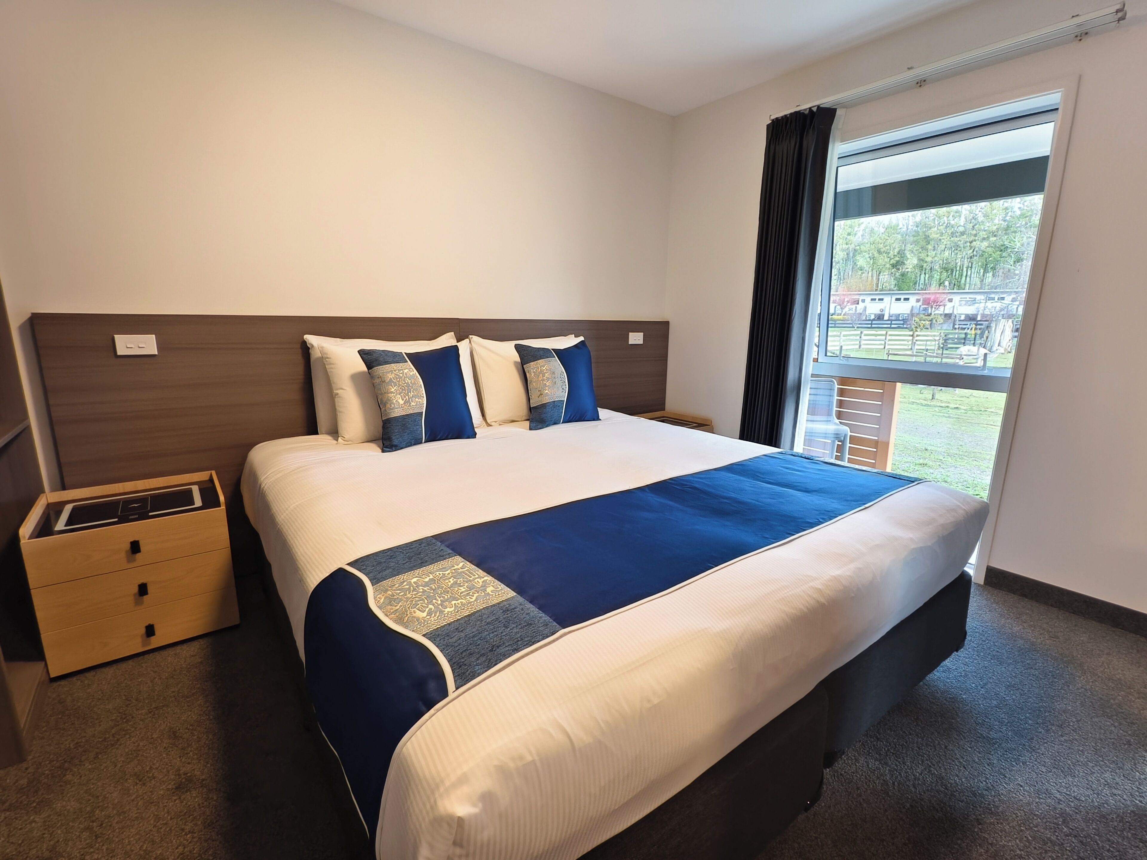 Quarto de hotel com uma cama grande, uma mesa de cabeceira, uma gaveta de madeira e uma janela com vista para o exterior.