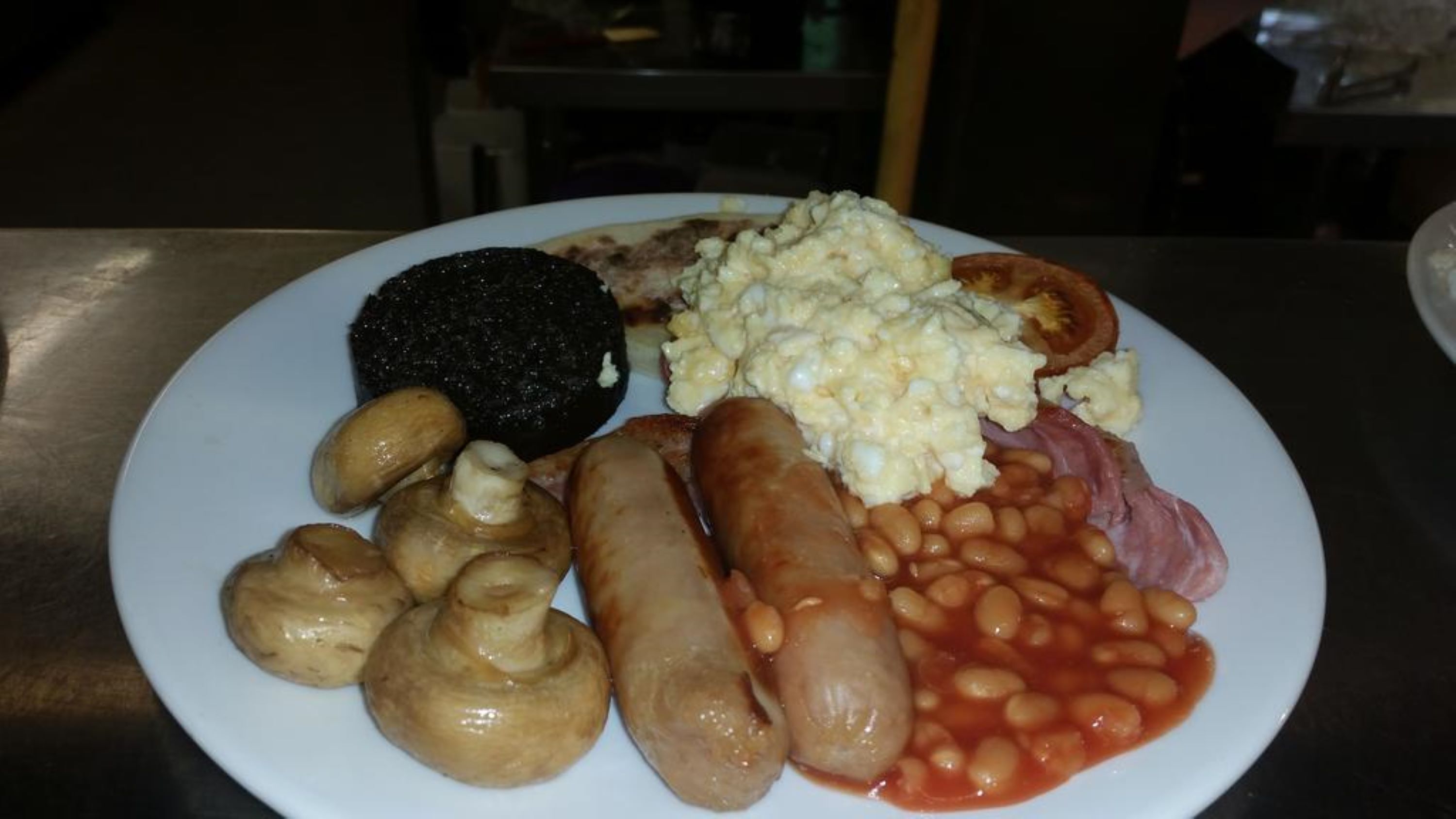 Um prato de comida com salsichas, feijão, cogumelos, ovos mexidos e um item preto.