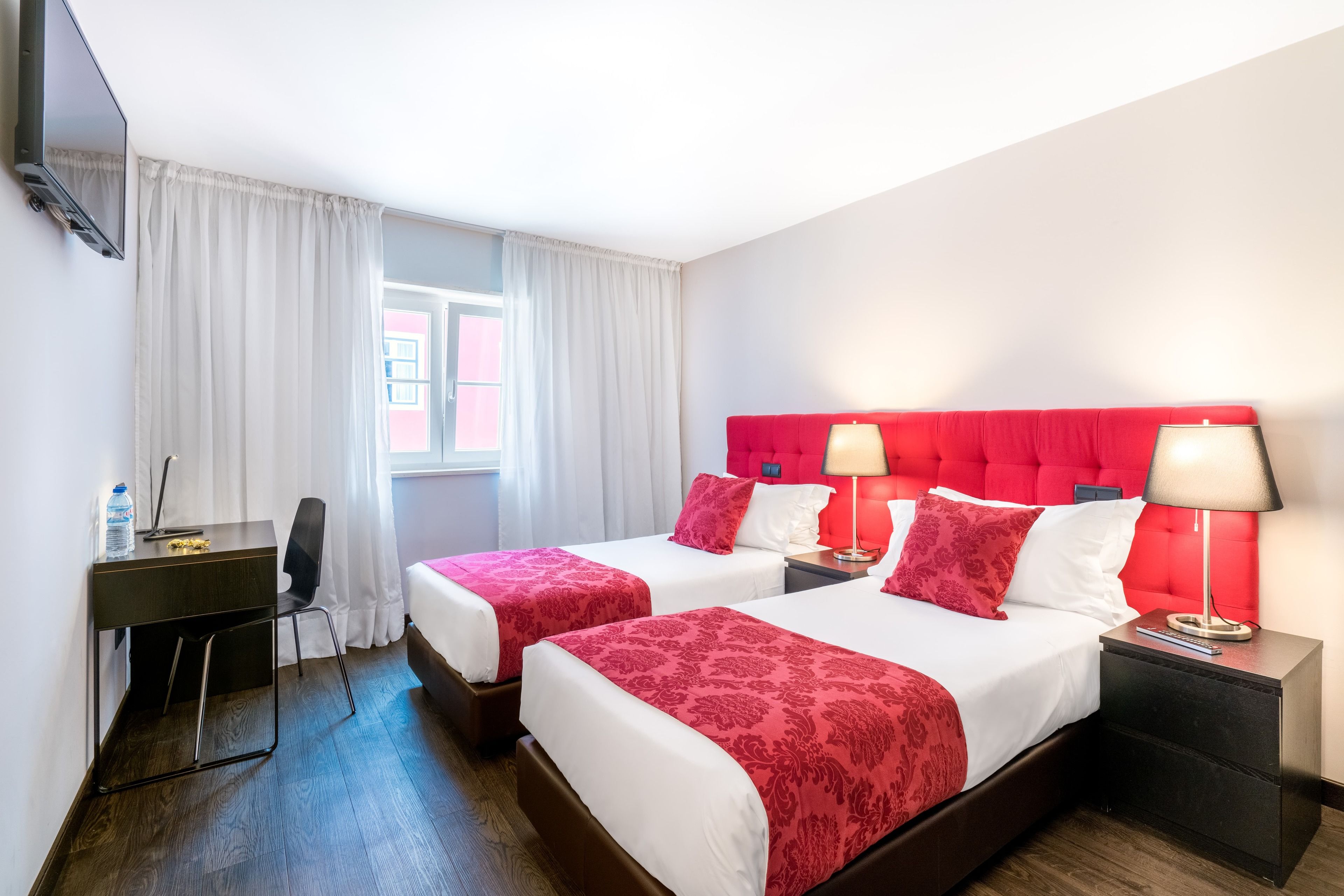 Quarto de hotel com duas camas, uma escrivaninha, uma cadeira, uma televisão e uma janela com cortinas.