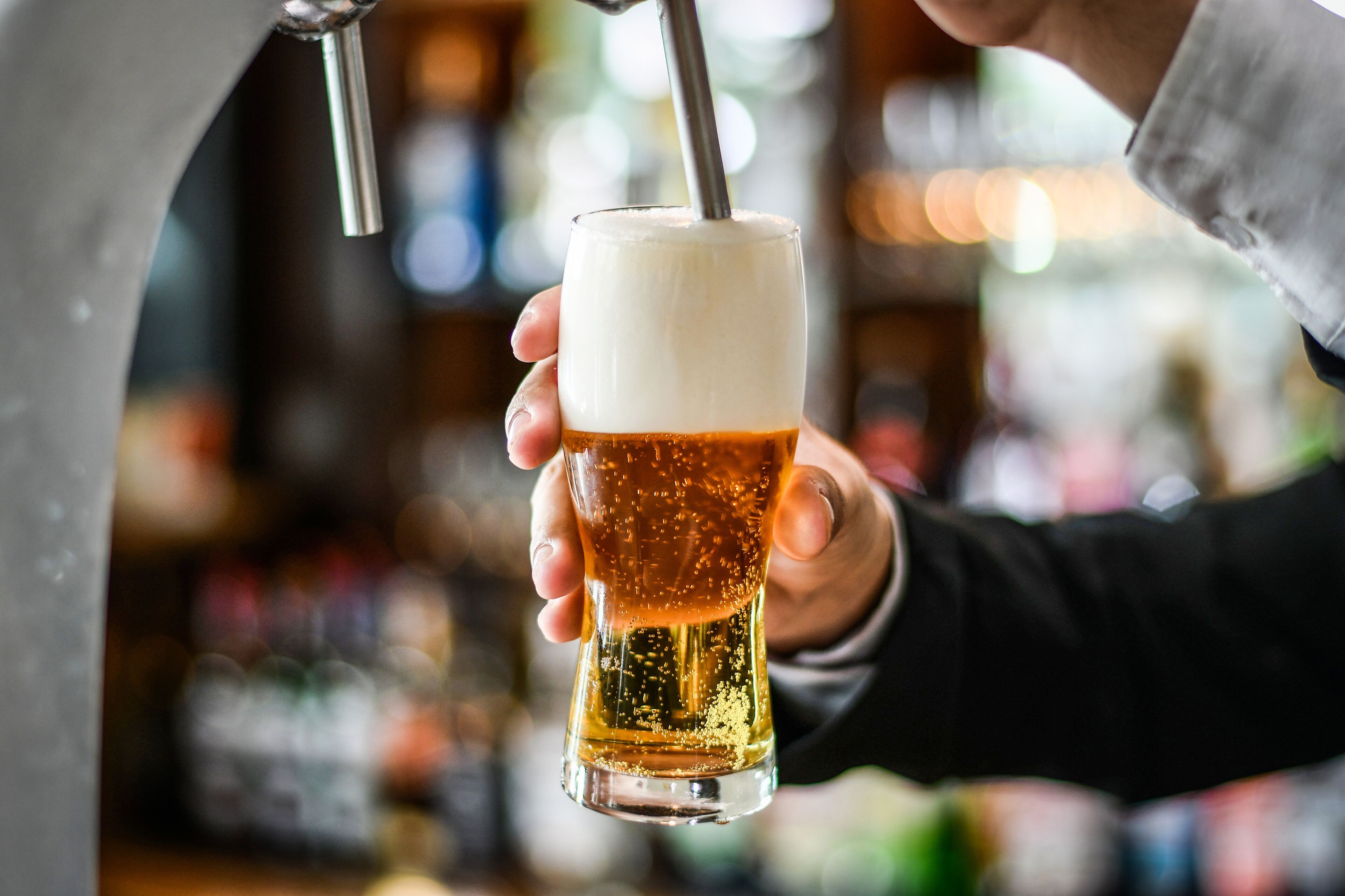 Uma pessoa segurando um copo de cerveja com canudo, com um fundo desfocado de prateleiras e garrafas.