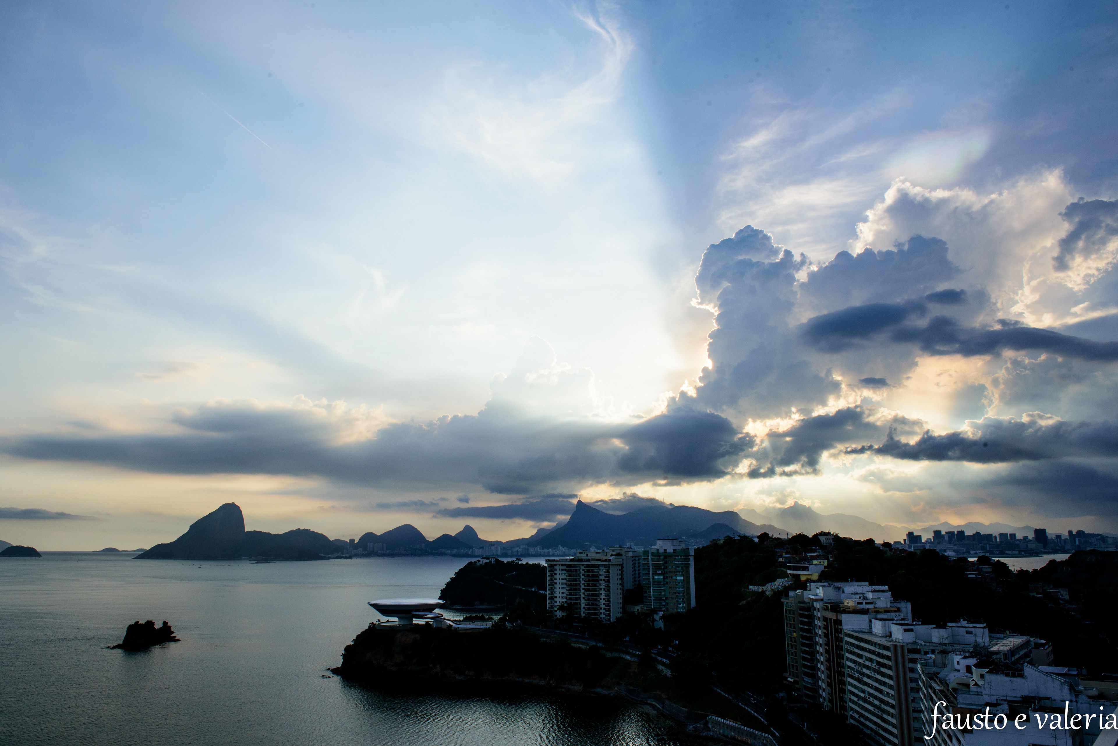 Paisagem costeira com edifícios próximos à água, uma montanha imponente ao fundo e céu parcialmente nublado.