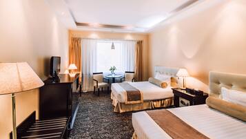 Quarto de hotel com cama, mesas de cabeceira, escrivaninha com televisão, uma cadeira, uma pequena mesa com uma planta e uma janela com cortinas.