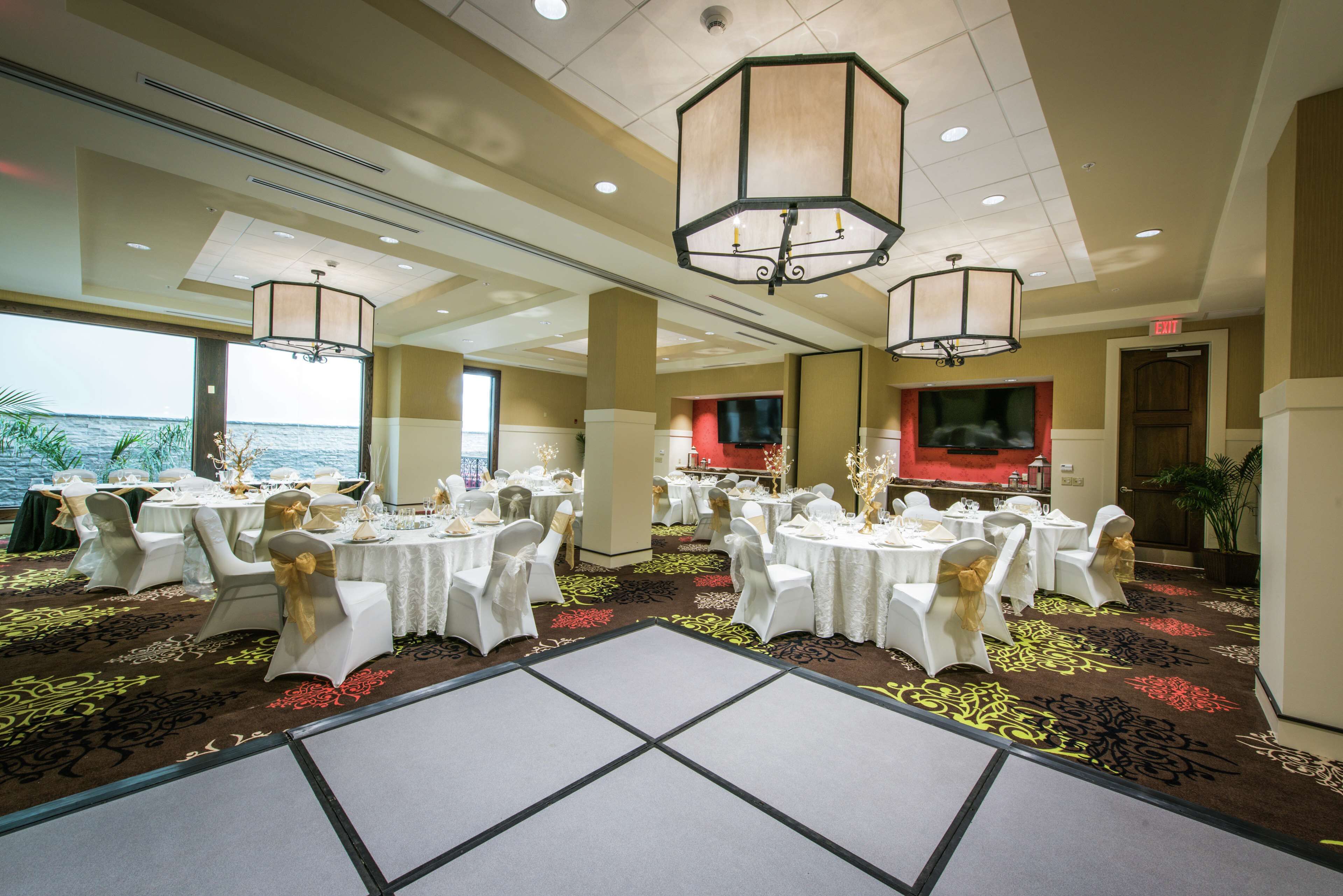 Uma sala de banquetes com mesas redondas, cadeiras brancas e fitas douradas, com uma grande janela e vista para o mar.