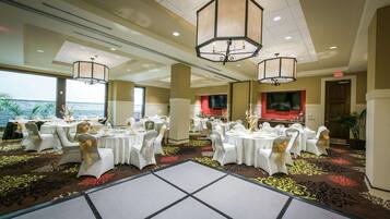 Uma sala de banquetes com mesas redondas, cadeiras brancas e fitas douradas, com uma grande janela e vista para o mar.
