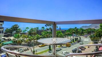 Varanda com mesa e cadeiras, vista para uma rua movimentada com palmeiras e o oceano.