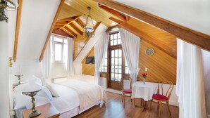 Quarto com teto inclinado, vigas de madeira, uma cama, uma mesa com cadeiras e uma janela com cortinas.