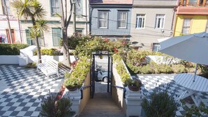 Escadaria que leva a um pátio com piso xadrez, rodeado por plantas em vasos e um banco.