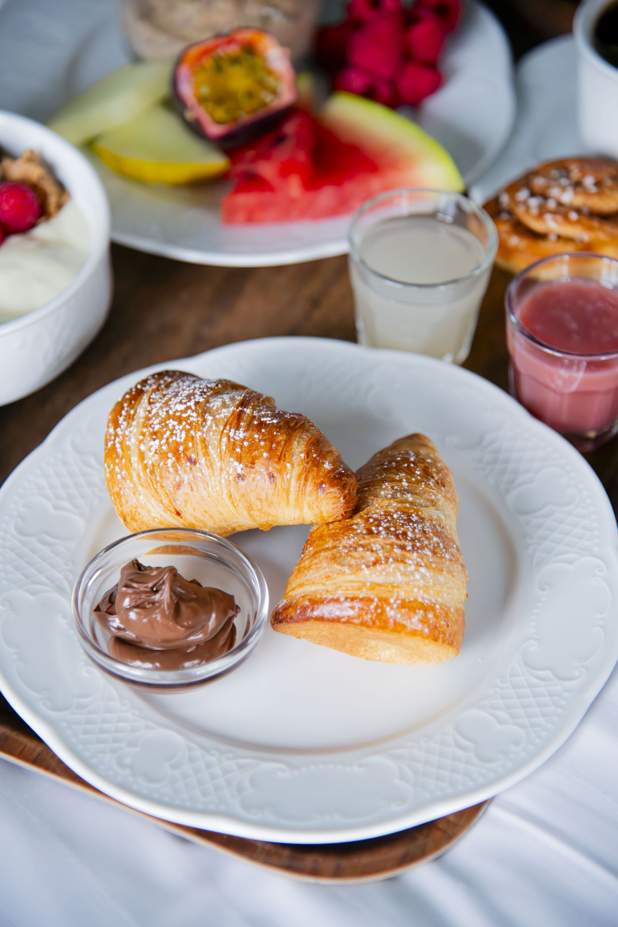Disposição para o pequeno-almoço com croissants, um pires de creme de chocolate e um prato com fruta e panquecas.