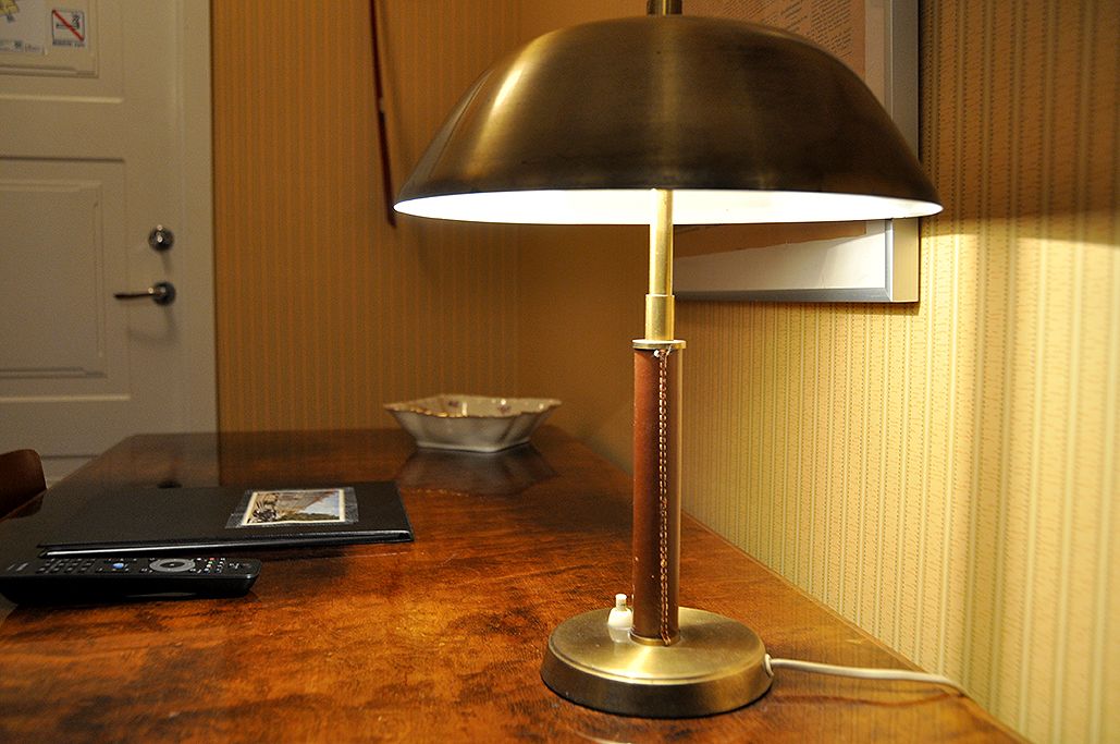 Uma mesa com uma luminária, um controle remoto e um laptop.