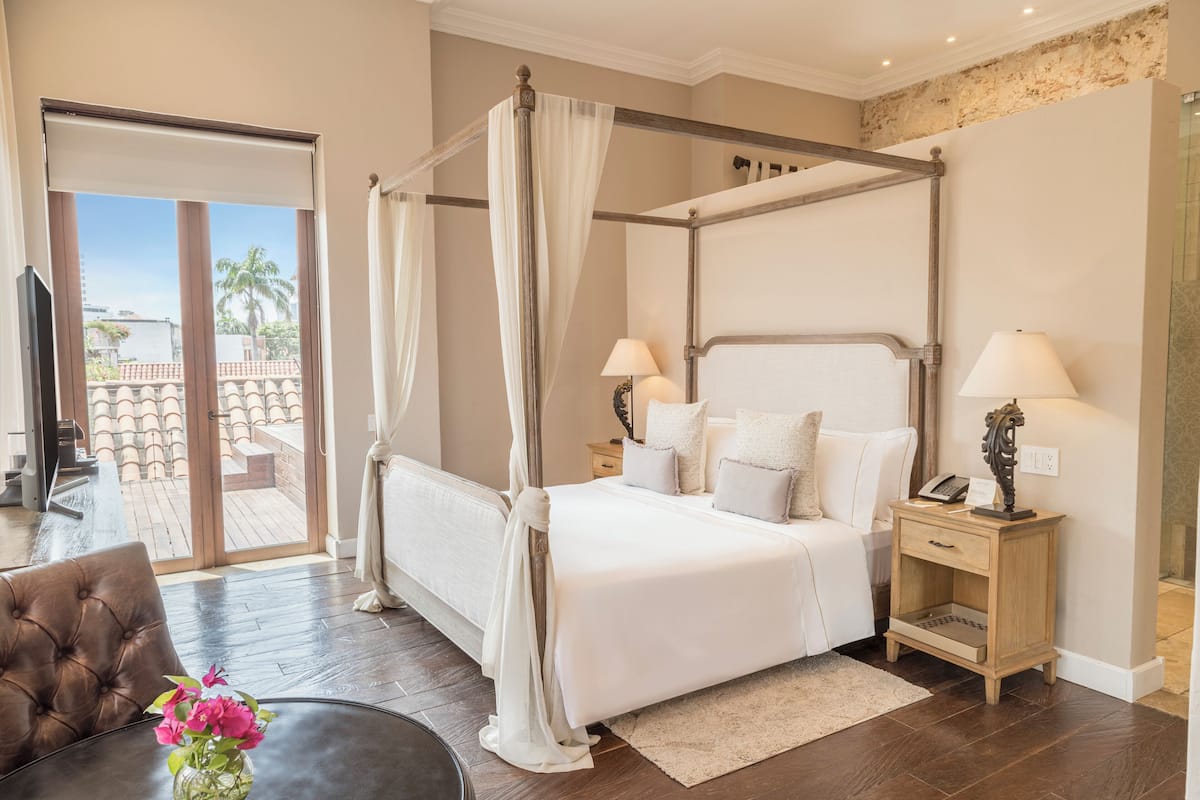 Quarto com cama de dossel, mesa de cabeceira com abajur e vista para a varanda.