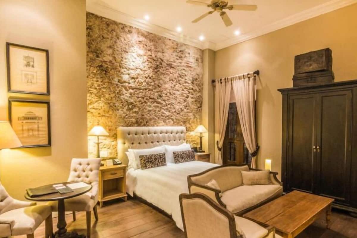 Quarto de hotel com parede de destaque em pedra, cama com lençóis brancos, mesa de cabeceira de madeira, escrivaninha com cadeira, sofá e armário de madeira escura.
