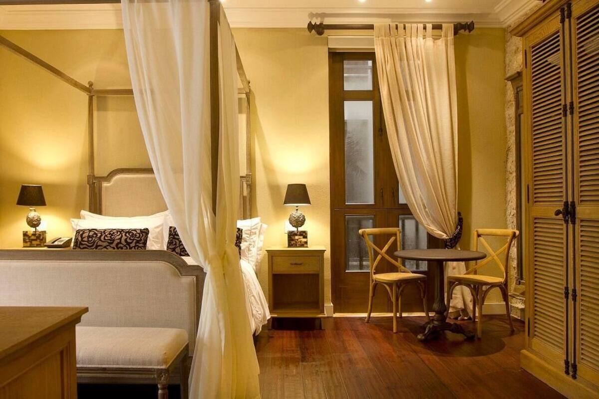 Quarto com cama de dossel, mesas de cabeceira com abajures, uma pequena mesa redonda com cadeiras e uma janela com cortinas.