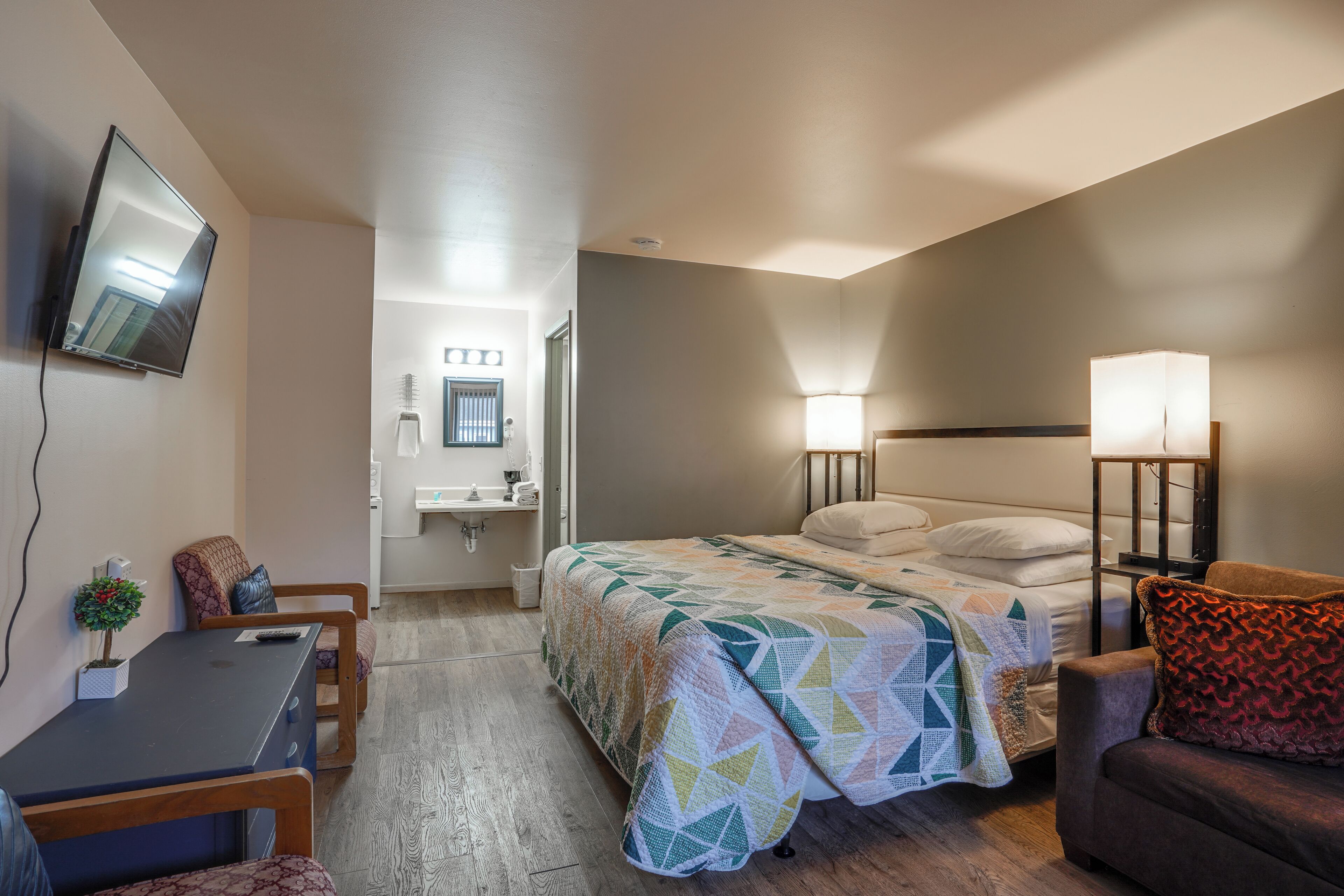 Quarto de hotel com cama, abajures, escrivaninha, cadeira e televisão presa na parede.